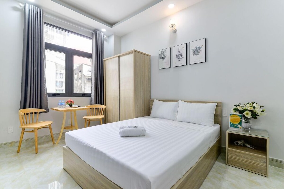 Quarto de hotel com cama, duas cadeiras, uma mesinha, guarda-roupa e mesa de cabeceira com um vaso de flores.
