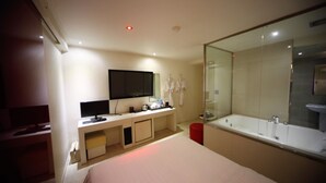 Quarto de hotel moderno com uma cama grande, televisão, secretária com computador, chuveiro em cabine de vidro e um cesto de lixo vermelho.