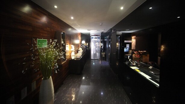 Um saguão de hotel com balcão de recepção, uma planta e um corredor que leva à entrada.