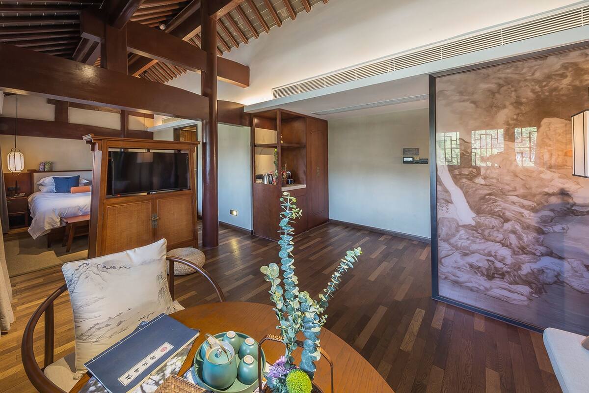 Quarto de hotel com piso de madeira, cama com televisão, área de refeições com mesa e cadeiras, e uma janela grande com vista para árvores.