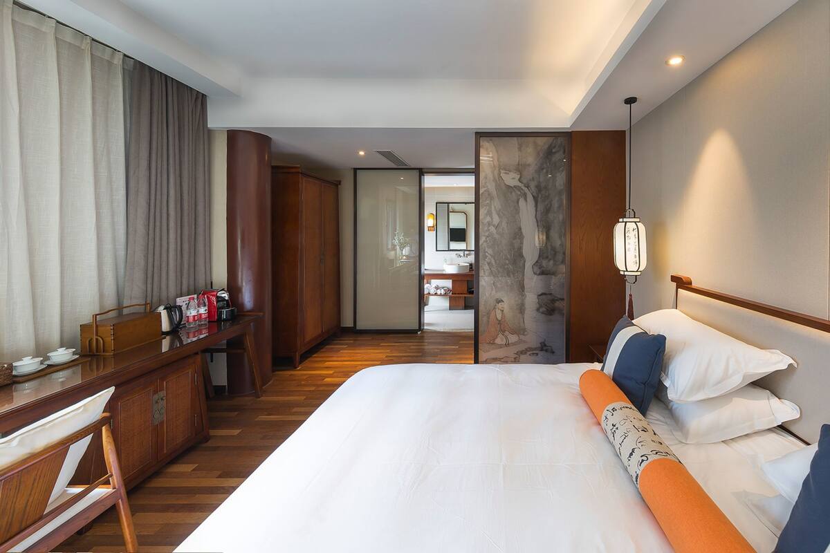 Quarto de hotel com uma cama grande, mobiliário de madeira, uma escrivaninha e um banheiro visível por uma porta aberta.