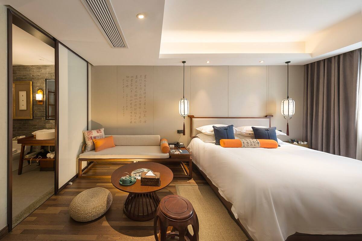Quarto de hotel moderno com uma cama grande, um sofá, uma pequena mesa redonda e um banheiro visível por uma porta aberta.