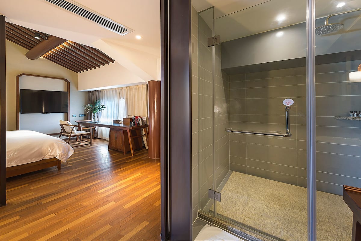 Quarto de hotel com piso de madeira, uma cama, uma escrivaninha com cadeira, área de chuveiro com porta de vidro e TV fixada na parede.