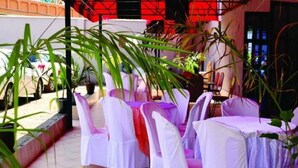 Um pátio externo com mesas e cadeiras cobertas por toalhas brancas e cor-de-rosa, rodeado por vasos com plantas.
