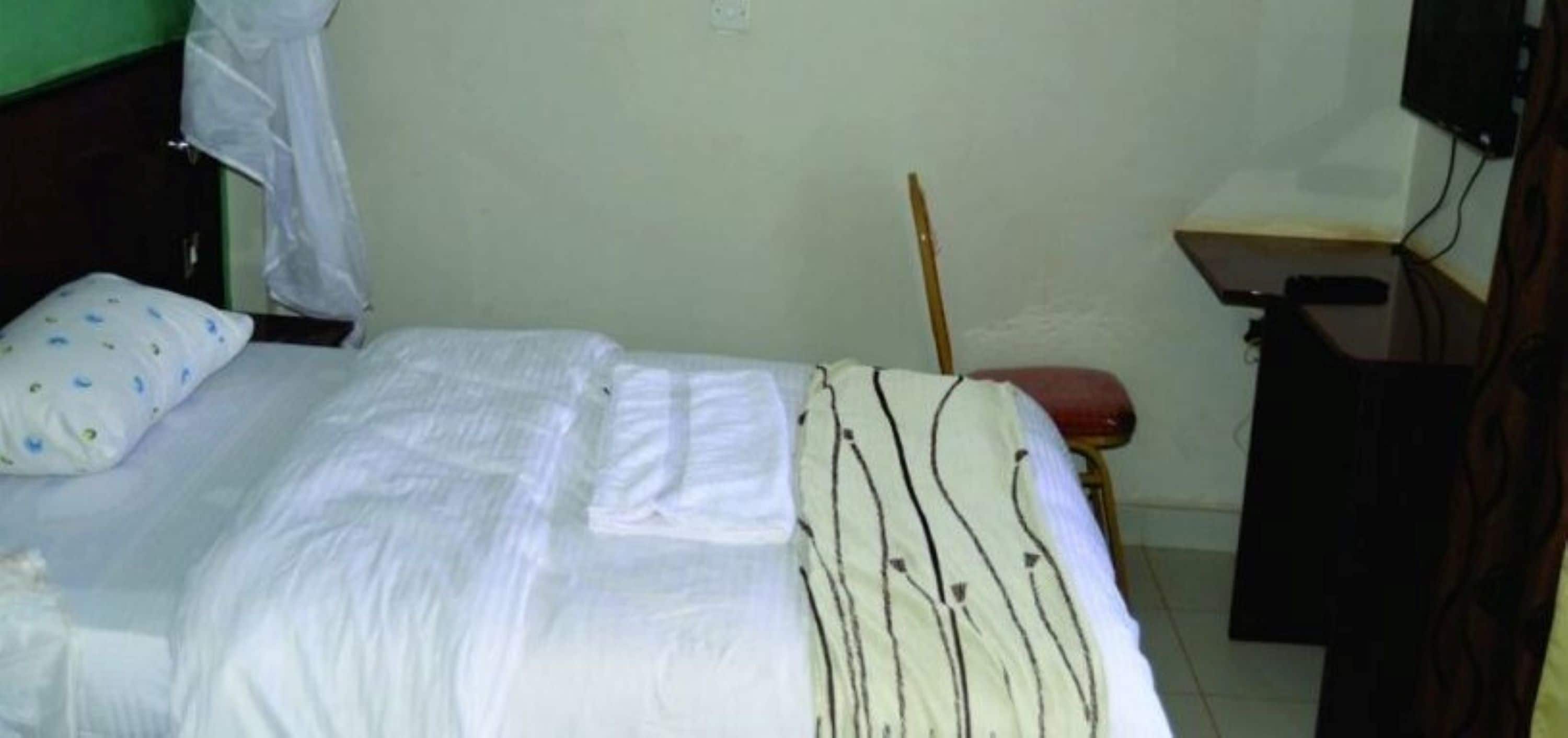 Uma cama de solteiro com roupa de cama branca e manta listrada, uma cadeira de madeira e uma pequena escrivaninha com cadeira.