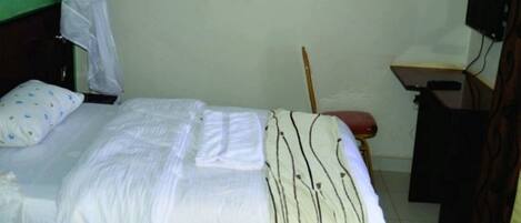 Uma cama de solteiro com roupa de cama branca e manta listrada, uma cadeira de madeira e uma pequena escrivaninha com cadeira.