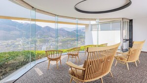 Quarto moderno com vista panorâmica de montanhas e cidade, mobiliado com cadeiras de madeira e mesa de vidro.