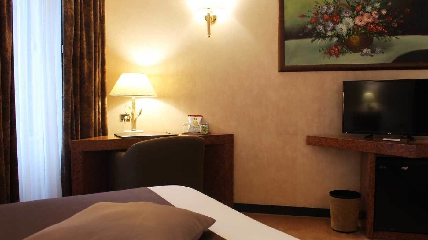 Quarto de hotel com uma cama, uma escrivaninha com abajur, televisão e um quadro na parede.