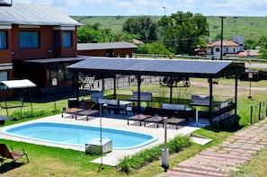 Piscina com espreguiçadeiras e área de estar coberta, cercada por um caminho de tijolos e vegetação.