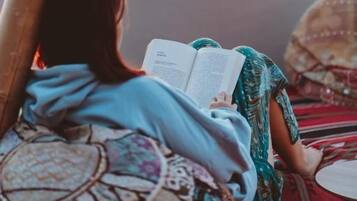 Uma pessoa sentada em um assento estampado, lendo um livro, com vista para um corpo d'água e colinas ao fundo.