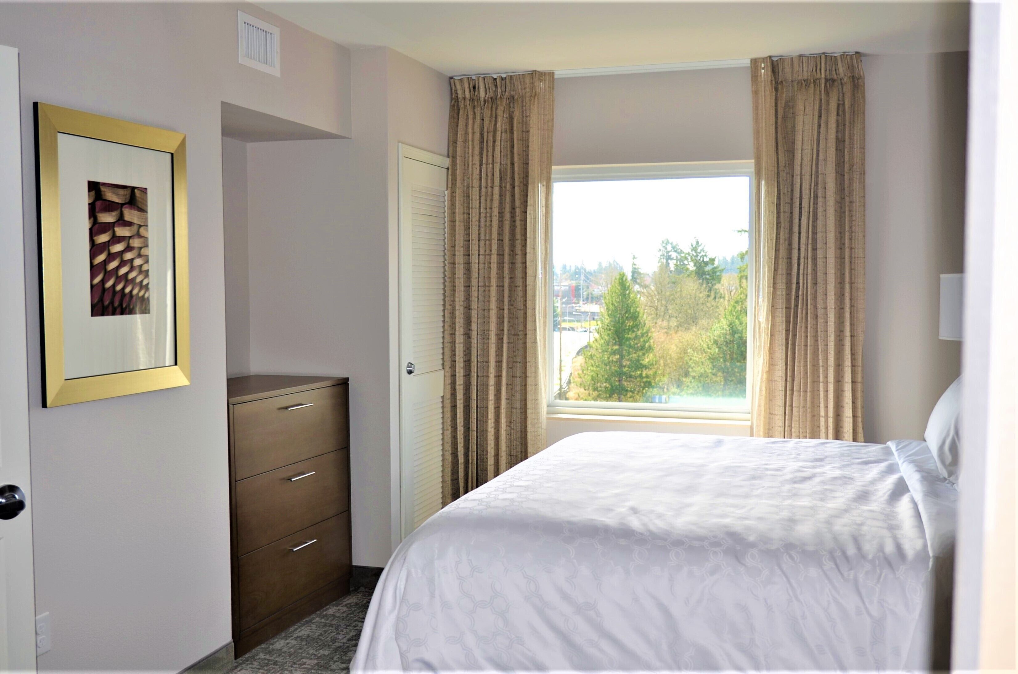 Quarto de hotel com uma cama, cômoda, quadro na parede e janela com cortinas.