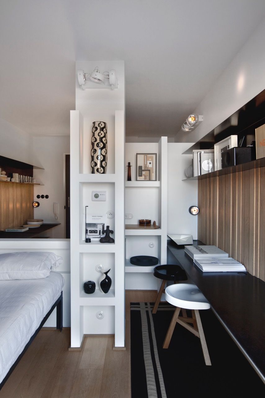 Quarto de hotel moderno com uma cama, uma estante branca com diversos objetos, uma pequena mesa com uma cadeira e uma parede de painéis de madeira.