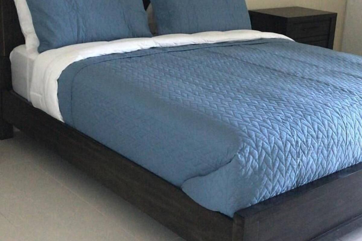 Uma cama com cabeceira escura, roupa de cama branca e azul, e um cobre-cama azul acolchoado.