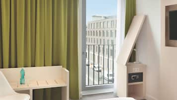 Quarto de hotel com cama, escrivaninha, cadeira, mesa de cabeceira com abajur, televisão e vista para um edifício através de uma janela.