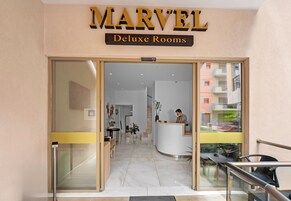 Entrada de hotel com balcão de recepção, porta de vidro e placa indicando 'MARVEL Deluxe Rooms'.