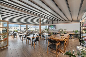 Um espaço amplo e interno com várias mesas e cadeiras, grandes janelas com vista para a cidade e teto com vigas aparentes.