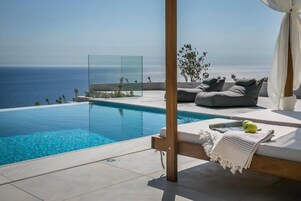 Uma piscina moderna infinita com vista desobstruída para o oceano, uma pérgola de madeira e um espaço de estar acolhedor com almofadas e manta.