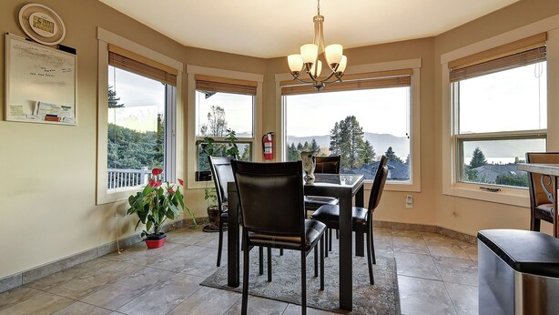 Uma sala de jantar com mesa e cadeiras, janelas amplas com vista para árvores e montanhas, um quadro branco na parede e um candelabro.