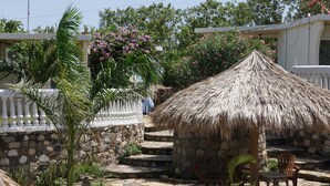 Uma cabana com telhado de palha, parede de pedra e degraus que levam até ela, rodeada por vegetação exuberante e plantas floridas.