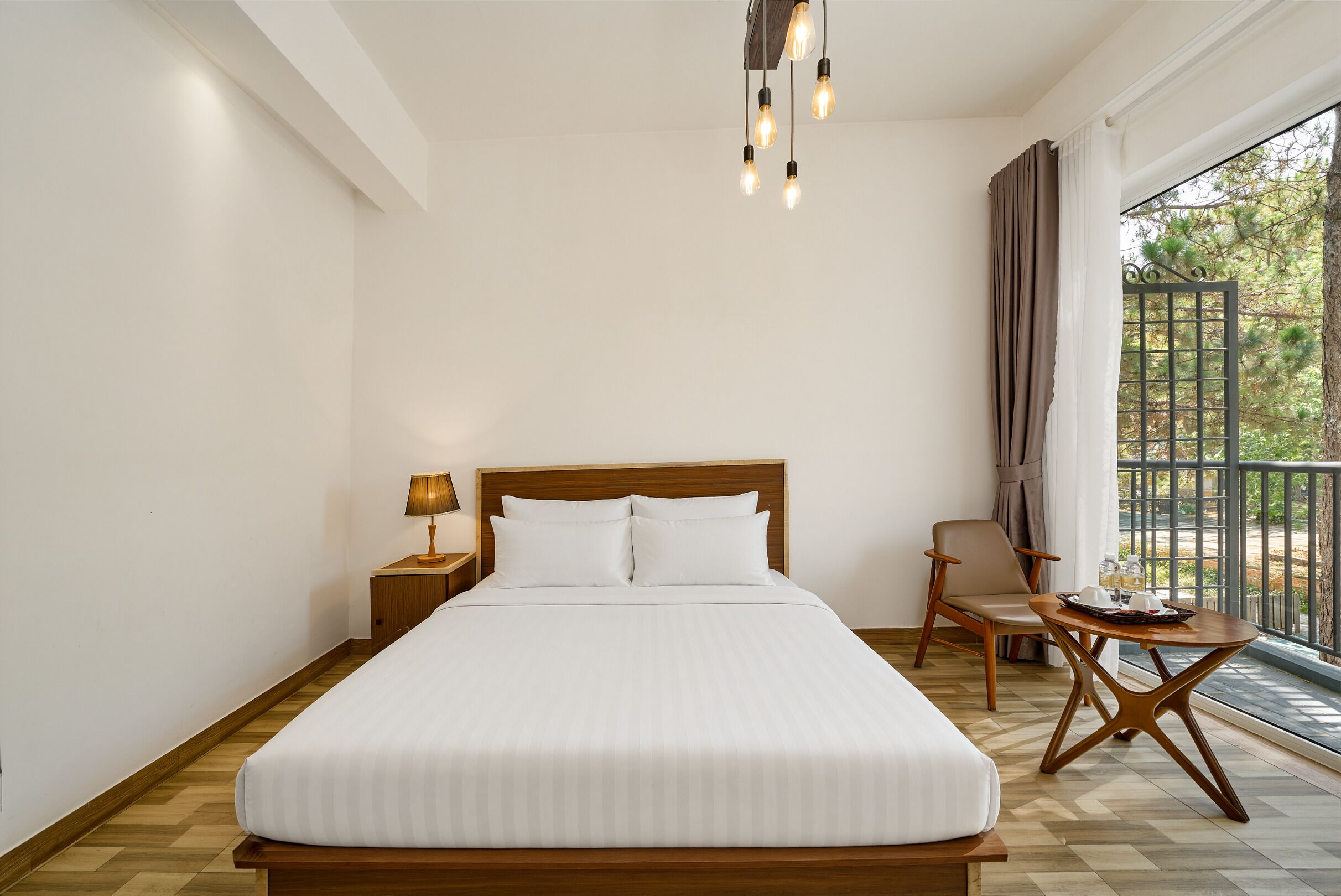 Quarto de hotel com uma cama grande, mesa de cabeceira com abajur, uma cadeira, pequena mesa com bandeja e varanda com vista.