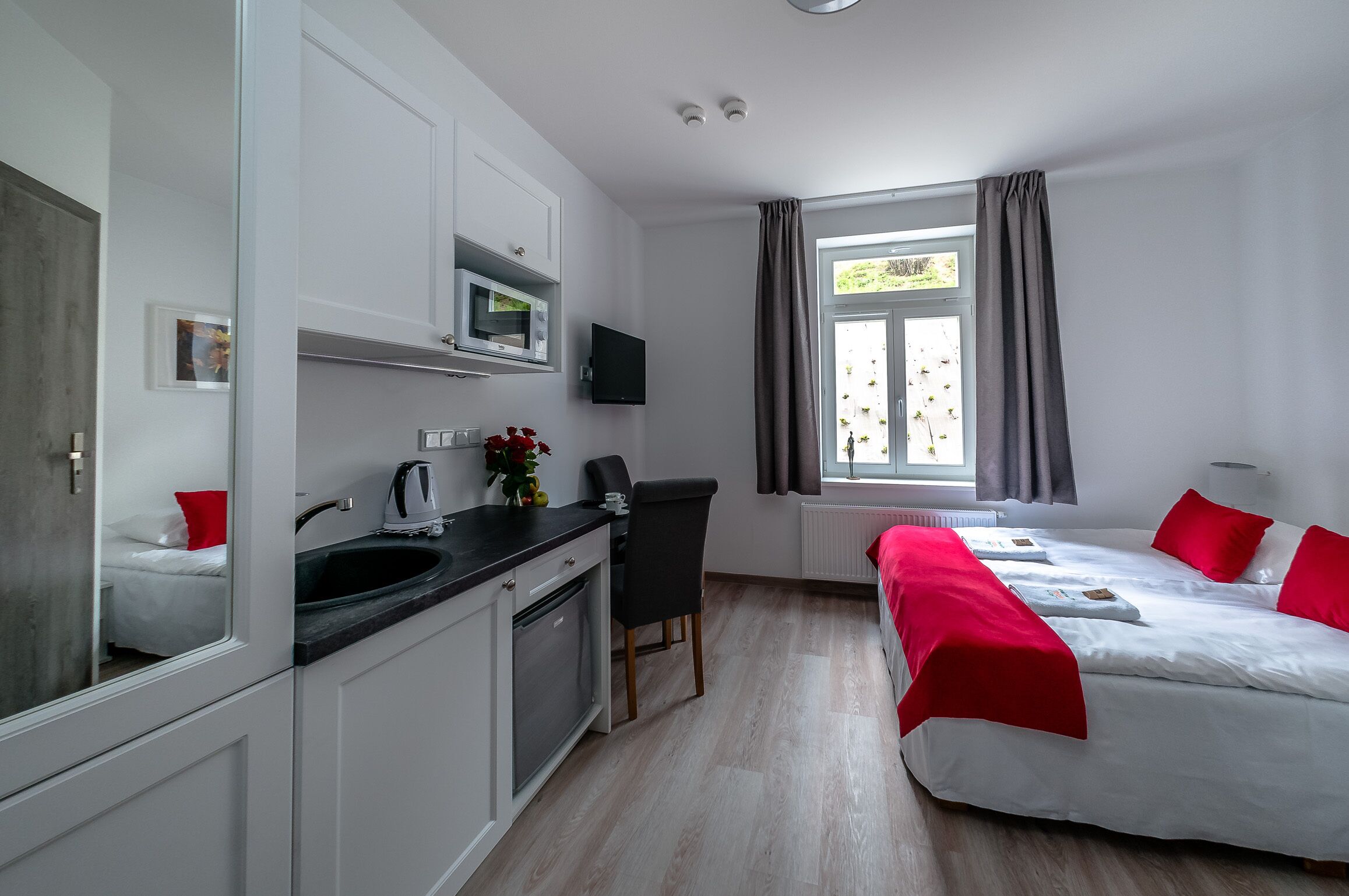 Quarto de hotel moderno com cozinha compacta, cama com roupa de cama vermelha, janela com cortinas e televisão de tela plana.