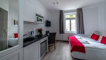 Quarto de hotel moderno com cozinha compacta, cama com roupa de cama vermelha, janela com cortinas e televisão de tela plana.