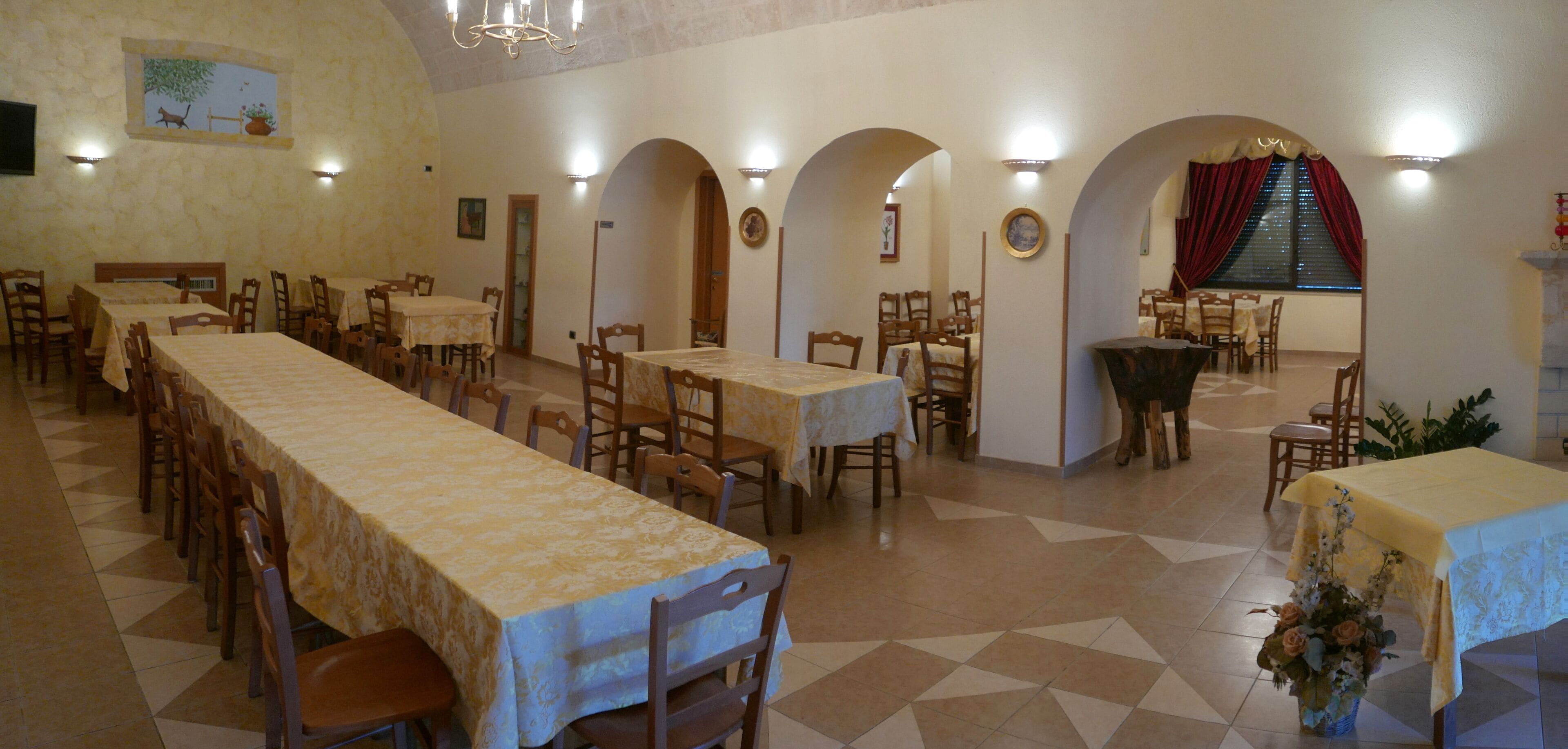 Uma sala de jantar com várias mesas e cadeiras, um lustre e uma janela com cortinas vermelhas.