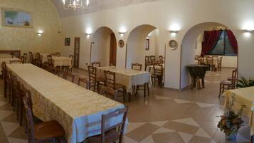 Uma sala de jantar com várias mesas e cadeiras, um lustre e uma janela com cortinas vermelhas.