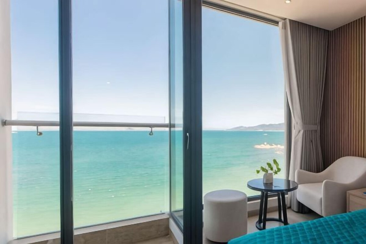 Quarto com uma grande janela com vista para a água, uma cama, uma cadeira e uma mesinha.