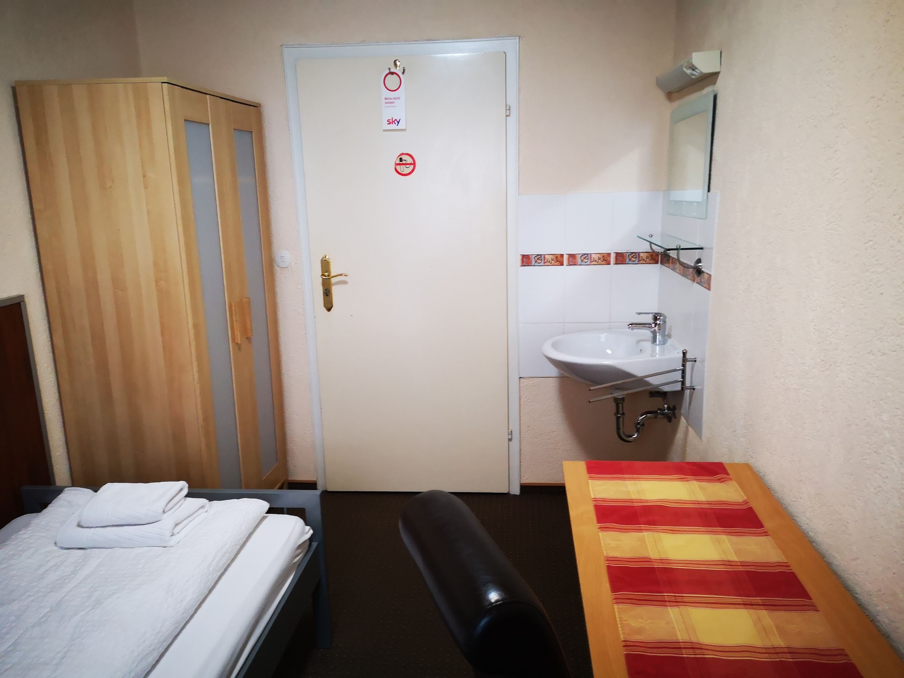 Quarto de hotel com cama, guarda-roupa de madeira, banheiro com pia e espelho, e uma mesa listrada.