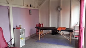 Um quarto com mesa de massagem, prateleira com objetos, uma cadeira vermelha e um ventilador.