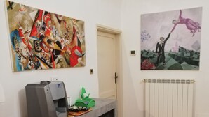 Um quarto com uma mesa consola cinzenta, uma impressora e uma planta verde. Há duas obras de arte nas paredes, uma abstrata e outra representando uma figura a alcançar um pássaro.