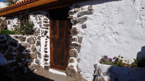 Um edifício rústico de pedra com porta de madeira e um pequeno jardim cheio de flores roxas.