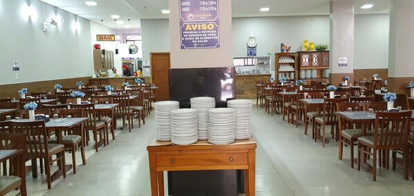 Restaurante com mesas e cadeiras de madeira, balcão de buffet e placa proibindo a venda de bebidas alcoólicas.