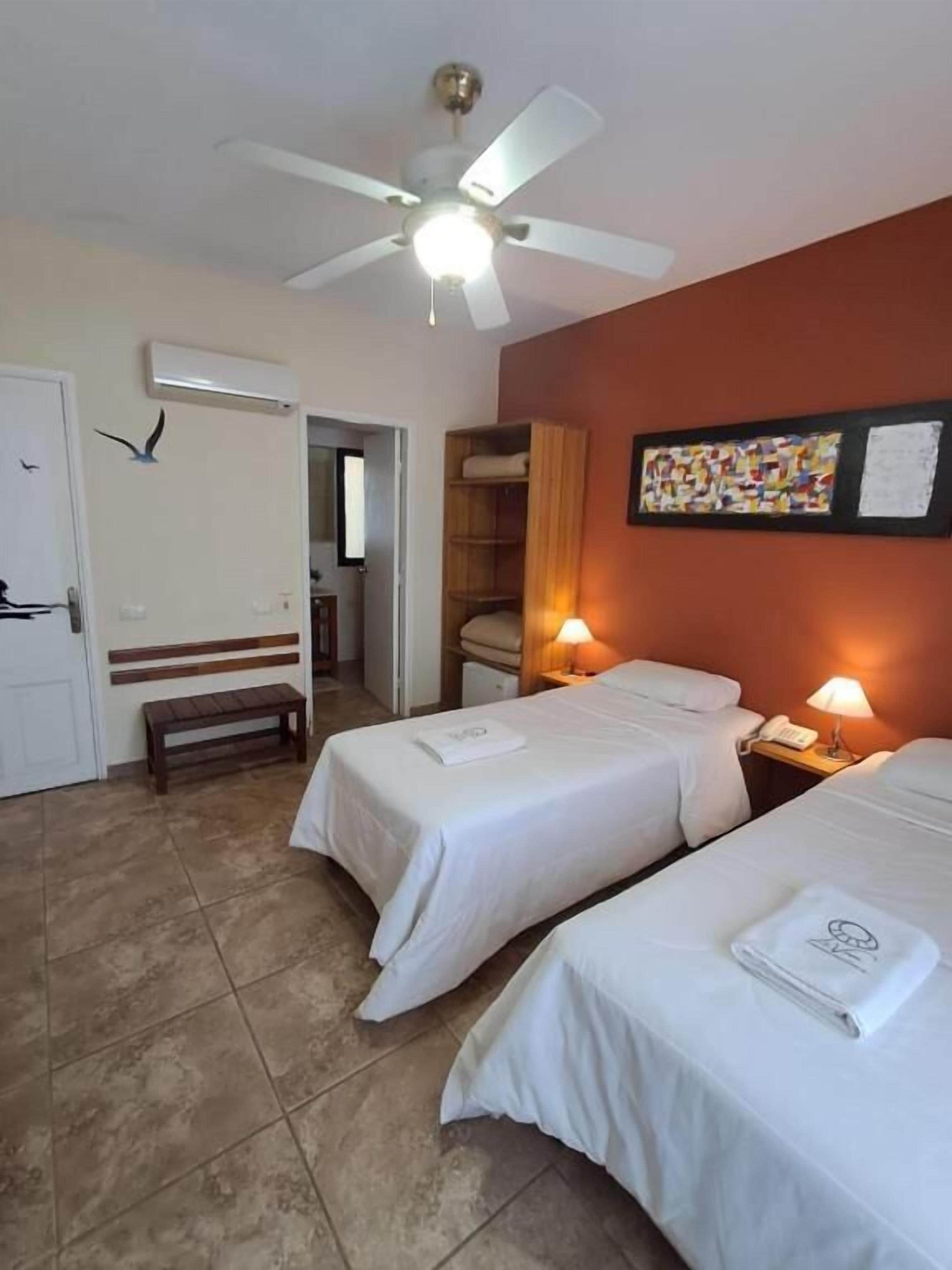 Quarto de hotel com duas camas, um ventilador de teto, um banco, uma escrivaninha e ar-condicionado fixado na parede.