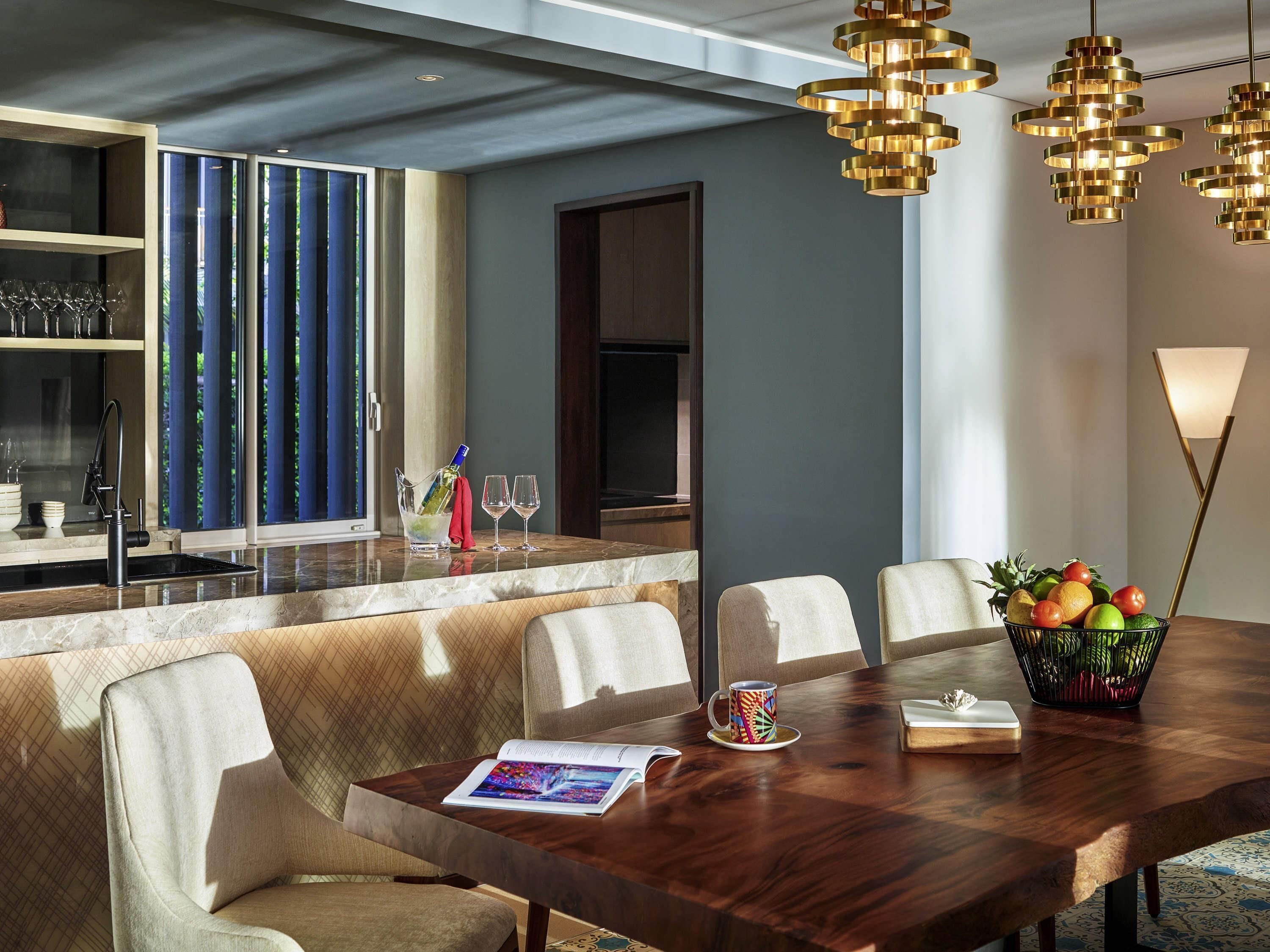 Uma sala de jantar moderna com mesa de madeira, cadeiras acolchoadas, conjunto de copos de vinho e uma cesta de frutas.