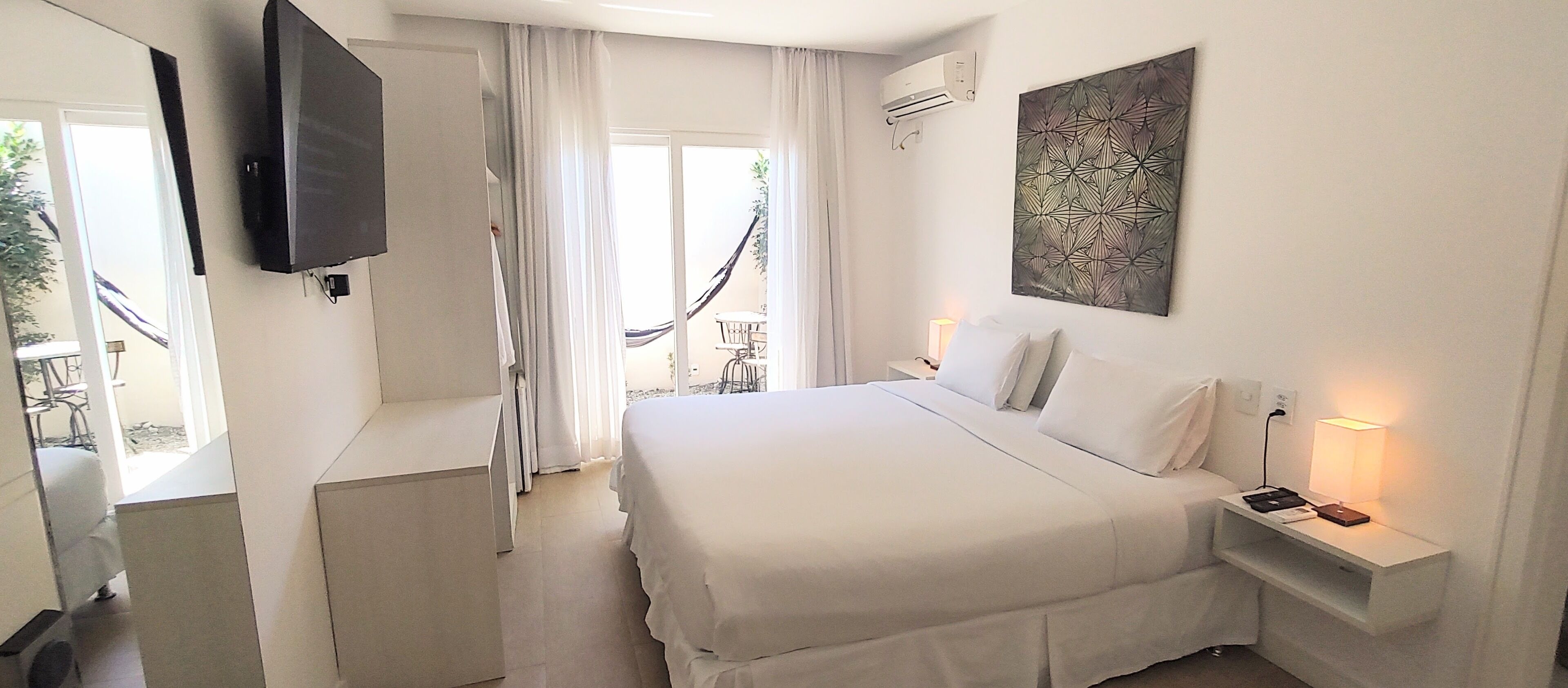 Quarto de hotel com cama, televisão fixada na parede, mesa de cabeceira com abajur e um quadro abstrato de grandes dimensões na parede.