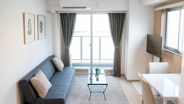 Uma sala de estar moderna com sofá, mesa de centro de vidro, televisão e grandes janelas com cortinas.