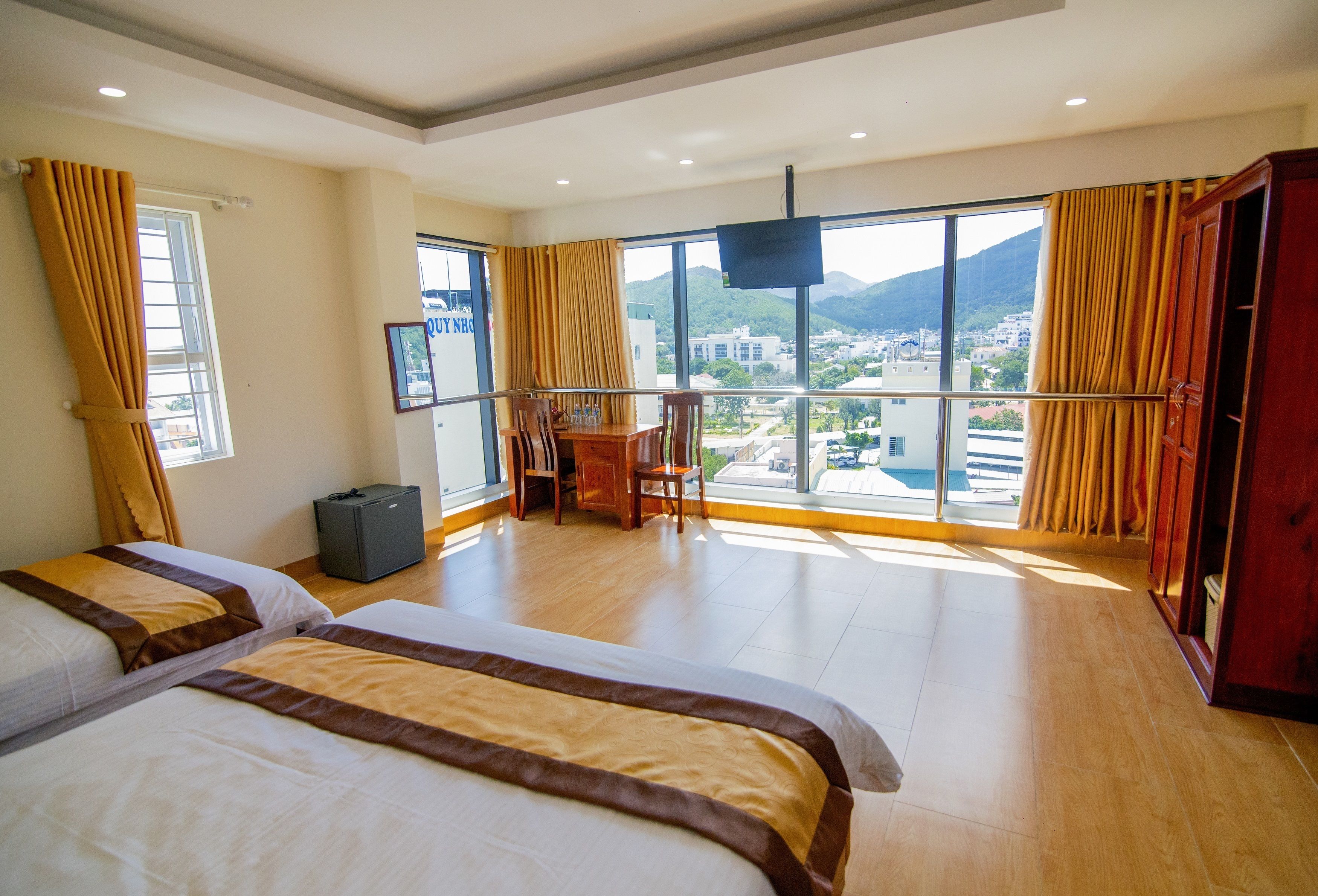 Quarto de hotel com duas camas, uma escrivaninha, uma cadeira, televisão e uma janela grande com vista para a cidade.