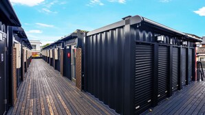 Um corredor com salas de metal preto, cada uma com uma porta entreaberta, que leva a um edifício com telhado de telhas.