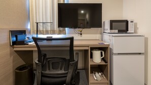 Quarto de hotel com escrivaninha, monitor de computador, micro-ondas, geladeira, cadeira, lixeira e prateleira com chaleira e um par de chinelos.