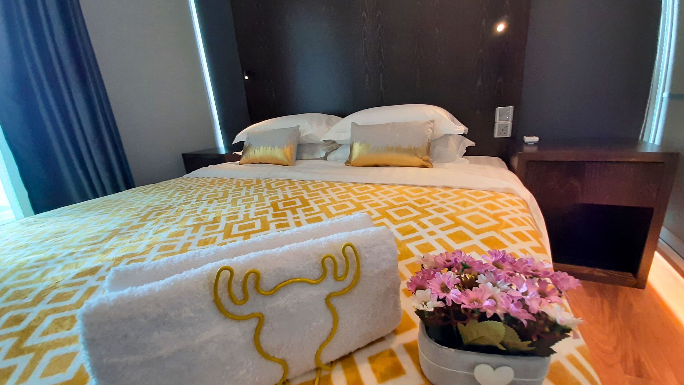 Uma cama bem arrumada com um espalha cama estampado, uma toalha branca com um desenho amarelo e uma planta em vaso com flores.
