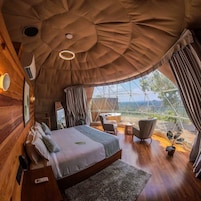 Quarto com cama grande, cabeceira de madeira, mesinha e uma cadeira. Há uma janela com cortinas e vista para o exterior.