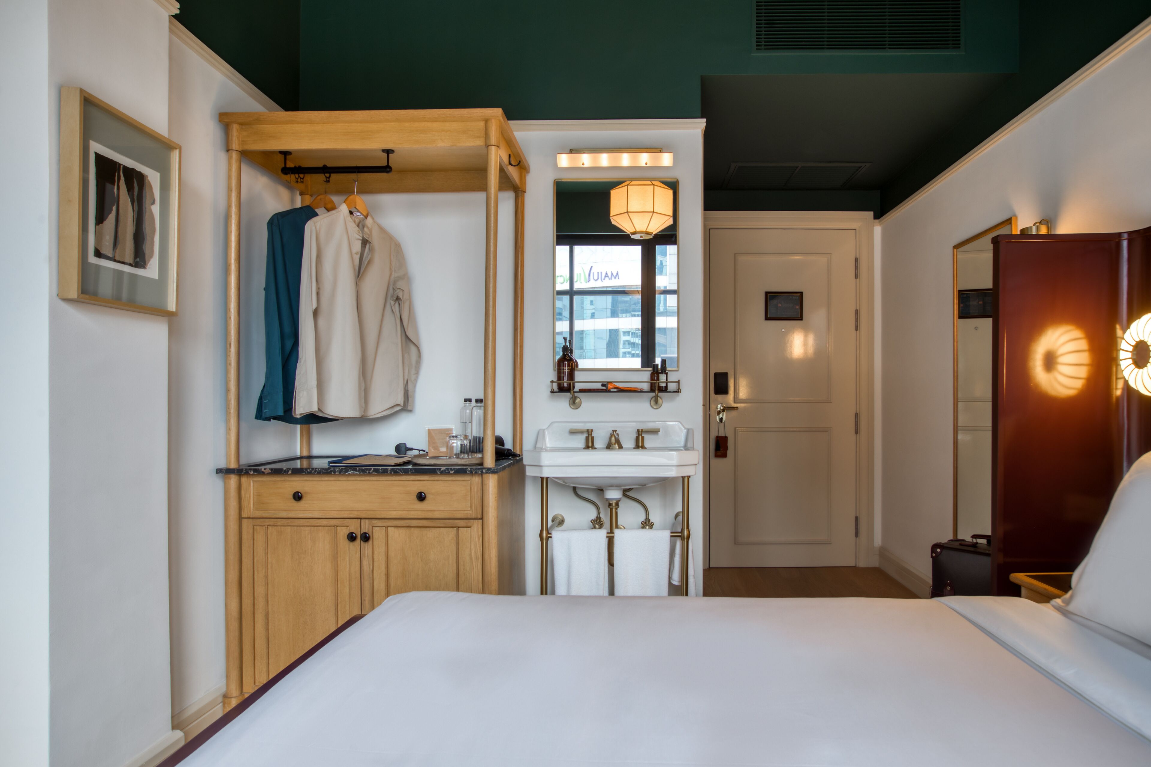 Quarto de hotel com cama, guarda-roupa de madeira, toucador embutido com espelho e janela com vista para a cidade.