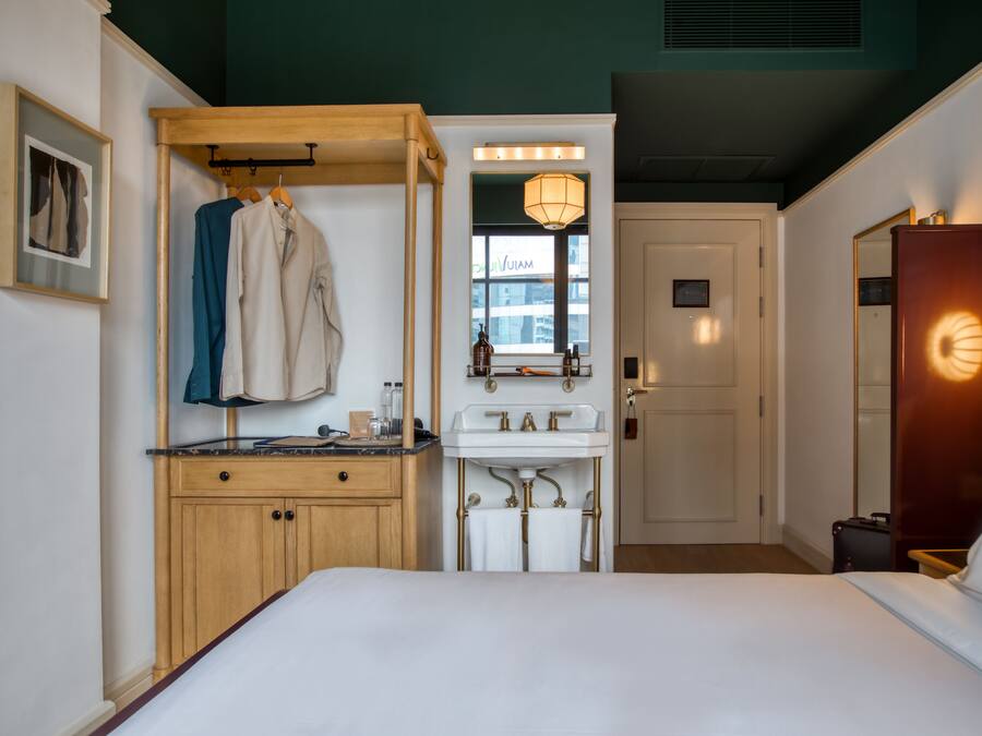 Quarto de hotel com cama, guarda-roupa de madeira, toucador embutido com espelho e janela com vista para a cidade.