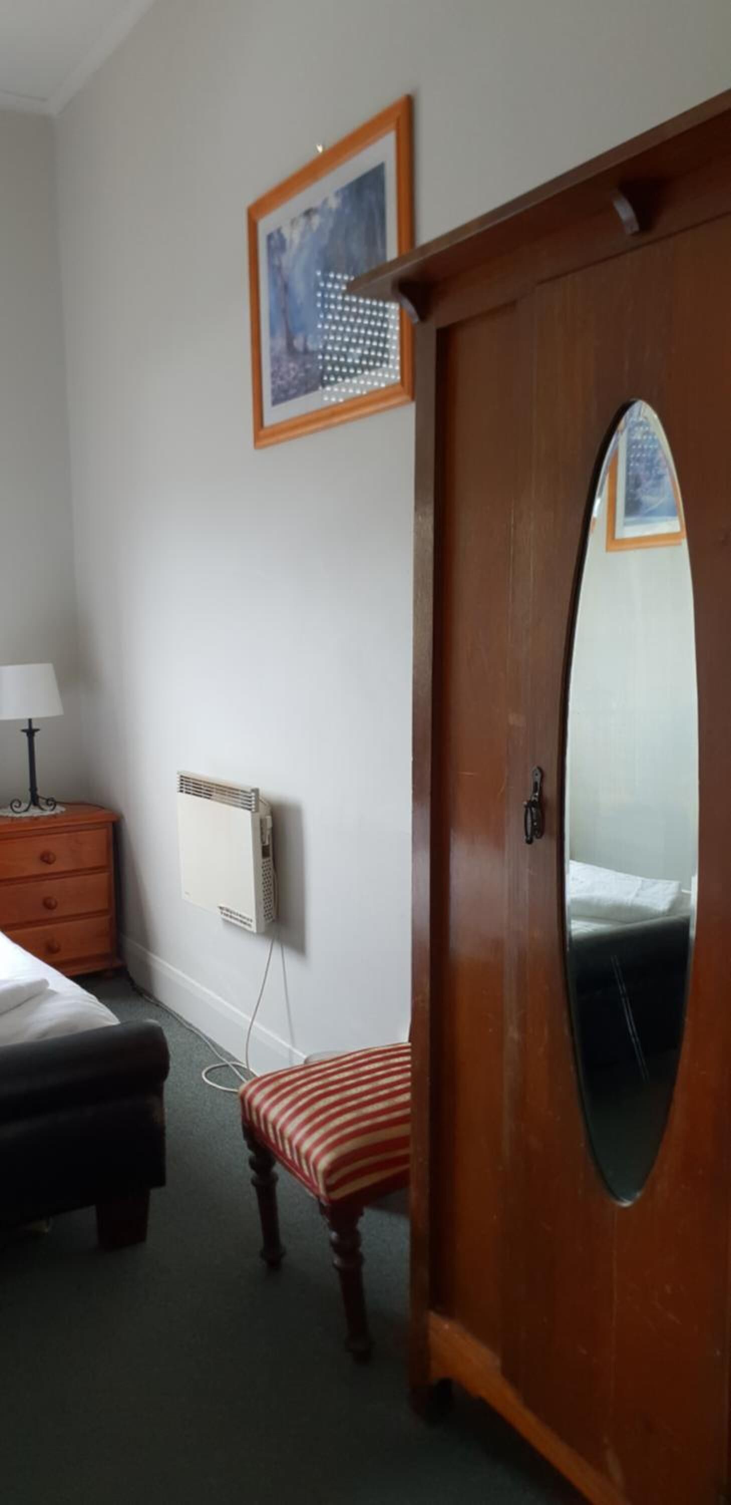 Quarto de hotel com guarda-roupa de madeira, uma cama, mesa de cabeceira com abajur, radiador e um quadro na parede.