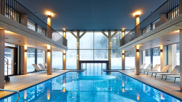 Uma grande piscina interior com deck de madeira, espreguiçadeiras e vista para a paisagem nevada através de amplas janelas.
