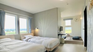 Quarto de hotel com duas camas, uma mesa de escritório com computador, uma cadeira, uma mesinha e vista para o exterior.
