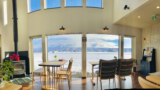 Quarto com uma grande janela com vista para uma paisagem nevada e um corpo d'água. Há uma mesa e cadeiras de madeira, uma pequena lareira e uma estante com livros.