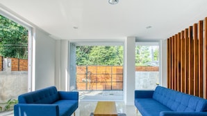Uma sala de estar moderna com dois sofás azuis, uma mesa de centro de madeira e janelas amplas que oferecem vista para uma parede de tijolos e vegetação lá fora.